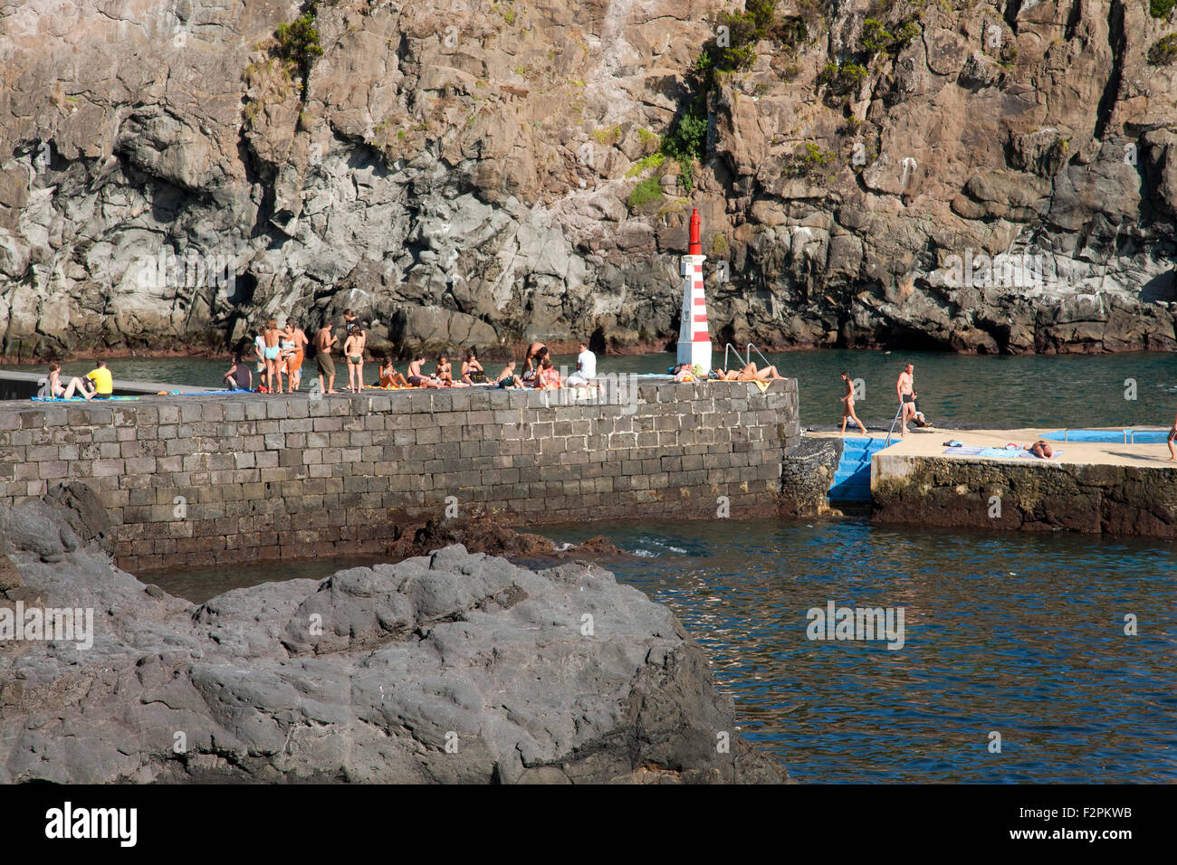People sunbathing in the harbour of Caloura surrounded by tall basalt ...