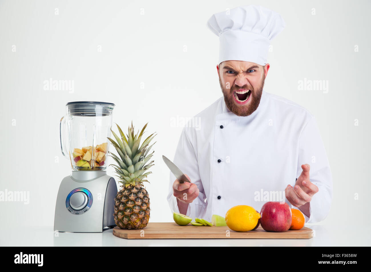Portrait of angry chef cook sitting at the table with fruits isolated ...