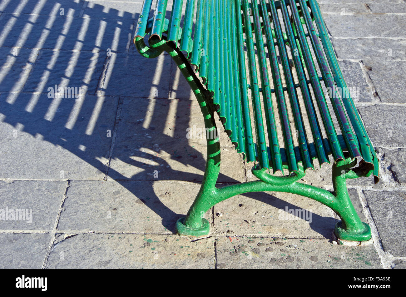 Sunlit green metal bench casting a shadow on pavement Stock Photo - Alamy