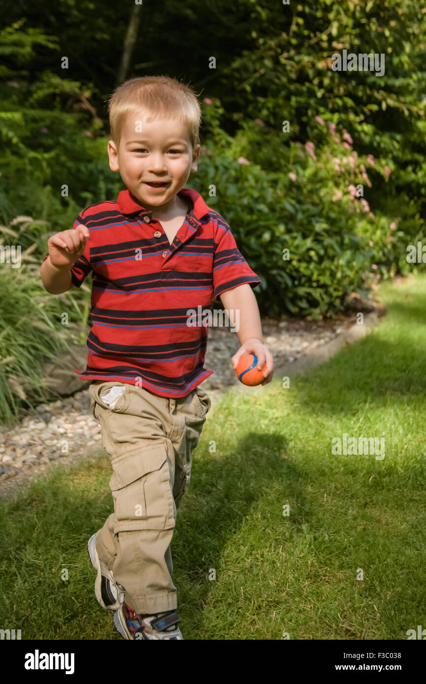 Two year old boy running with a small ball in his hand in the backyard ...