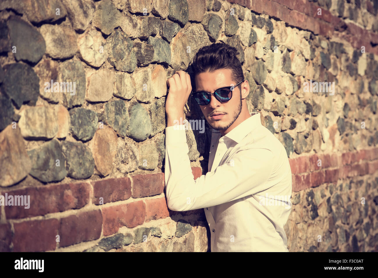Attractive young man sitting against brick wall, looking at camera ...