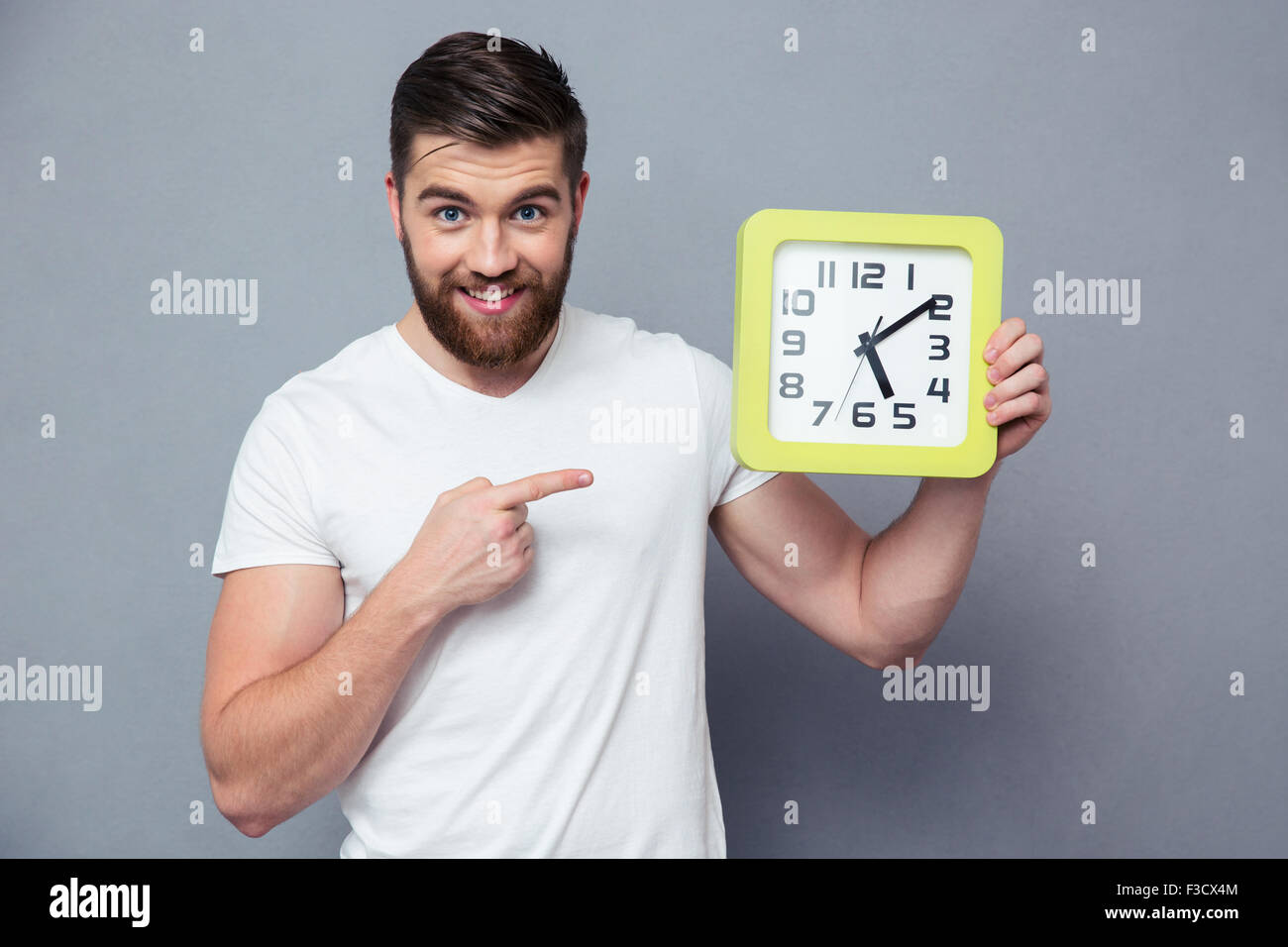 Portrait of a happy casual man pointing finger on wall clock over gray ...