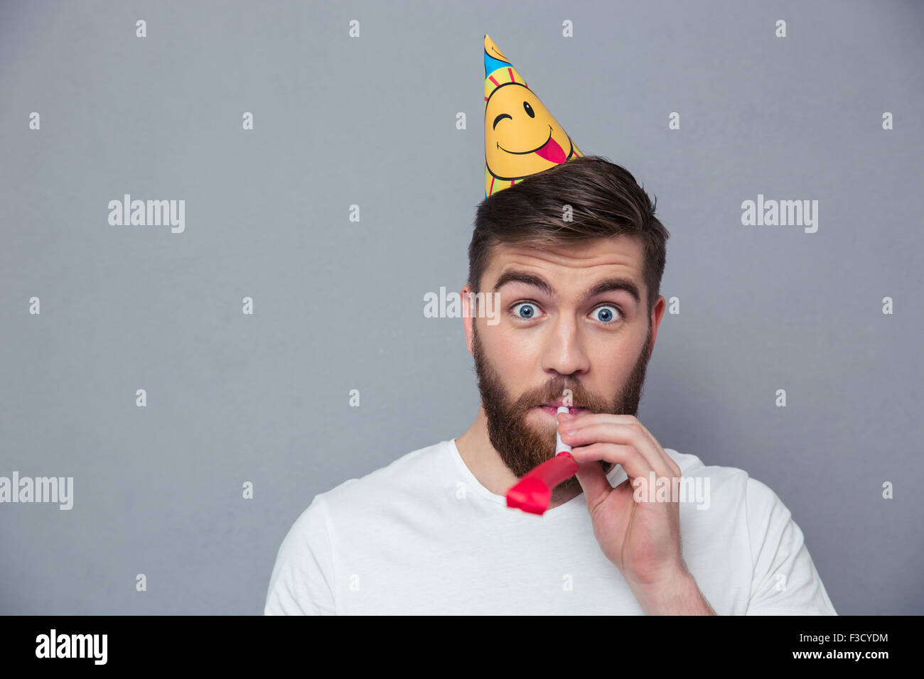 Portrait of a handsome man with party hat blowing in whistle over gray ...
