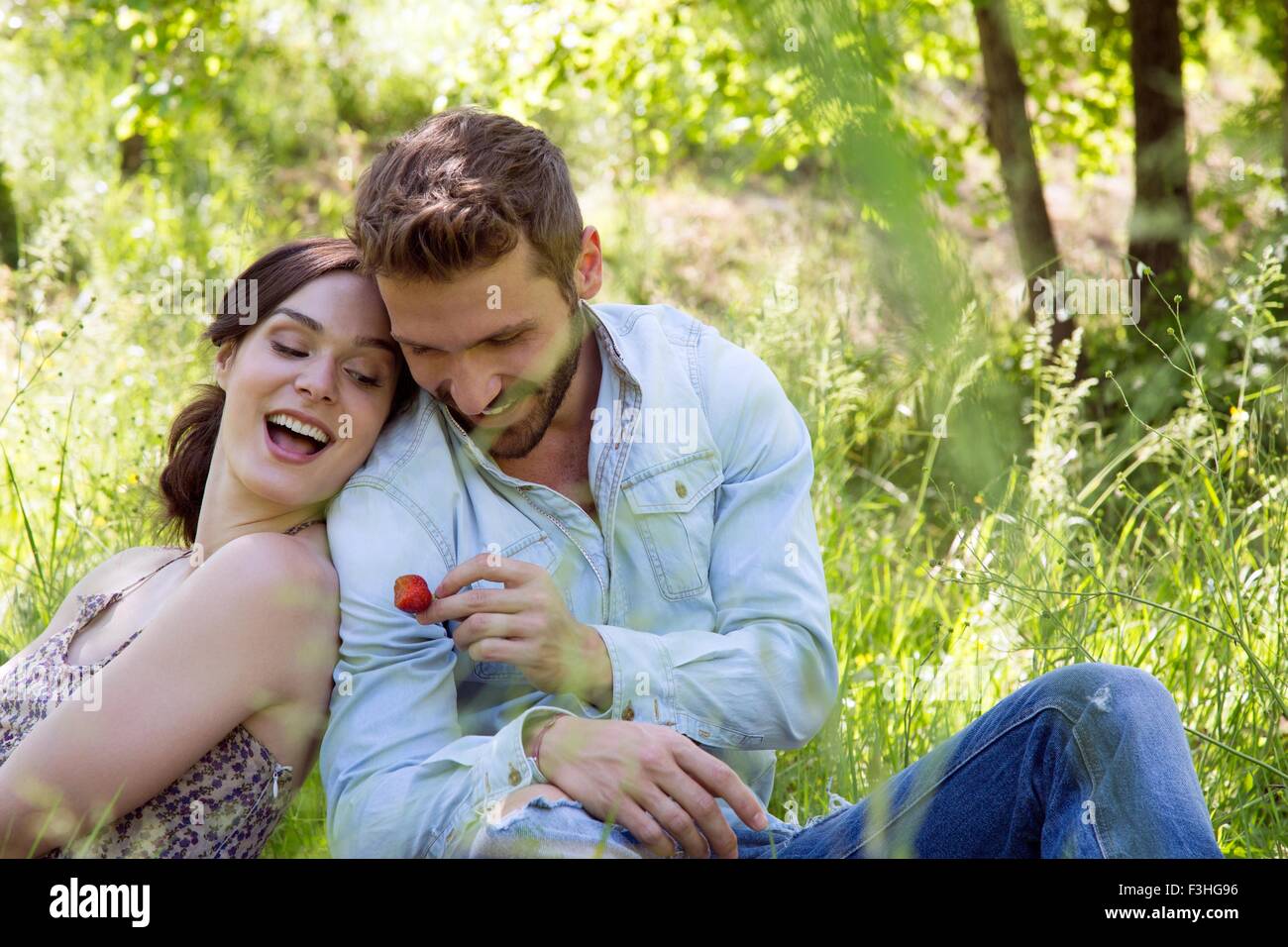 Young couple sitting back to back on grass holding strawberry smiling ...