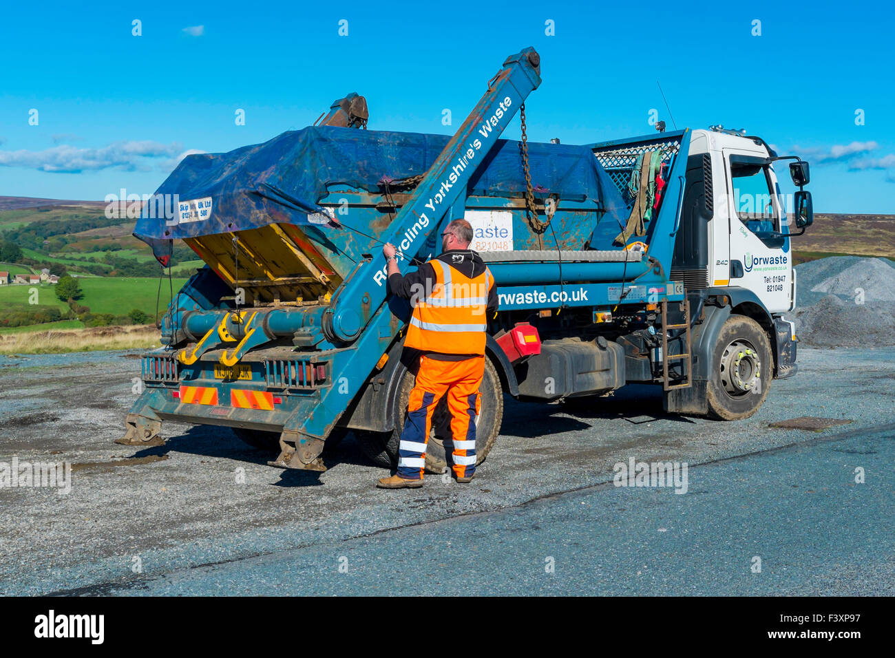 Driver securing tarpaulin cover over a waste skip on a truck beside a ...
