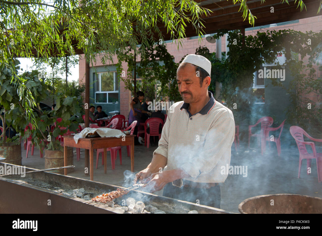 China, Xinjiang, Manas. A man cooks meat on an open pit barbecue at a ...