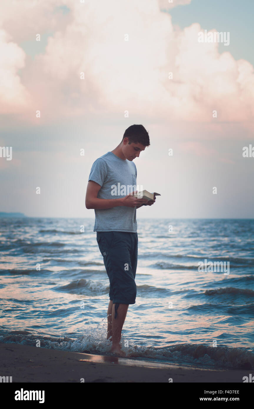 young man reading a book standing in the waves on a beach with the sun ...