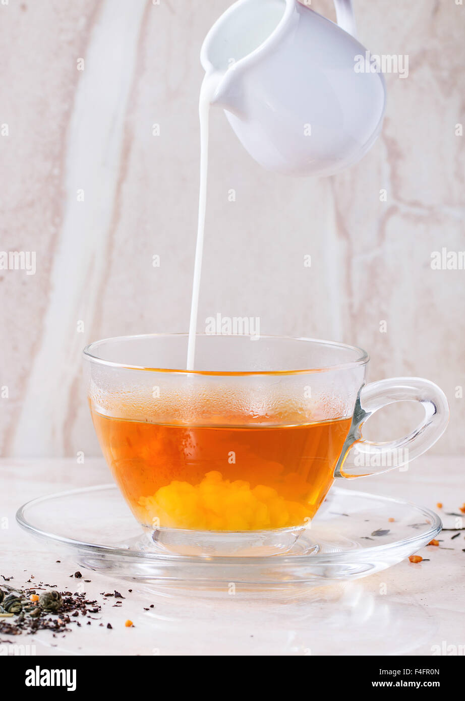 Milk pouring from white jug to glass cup of hot tea on saucer with dry ...