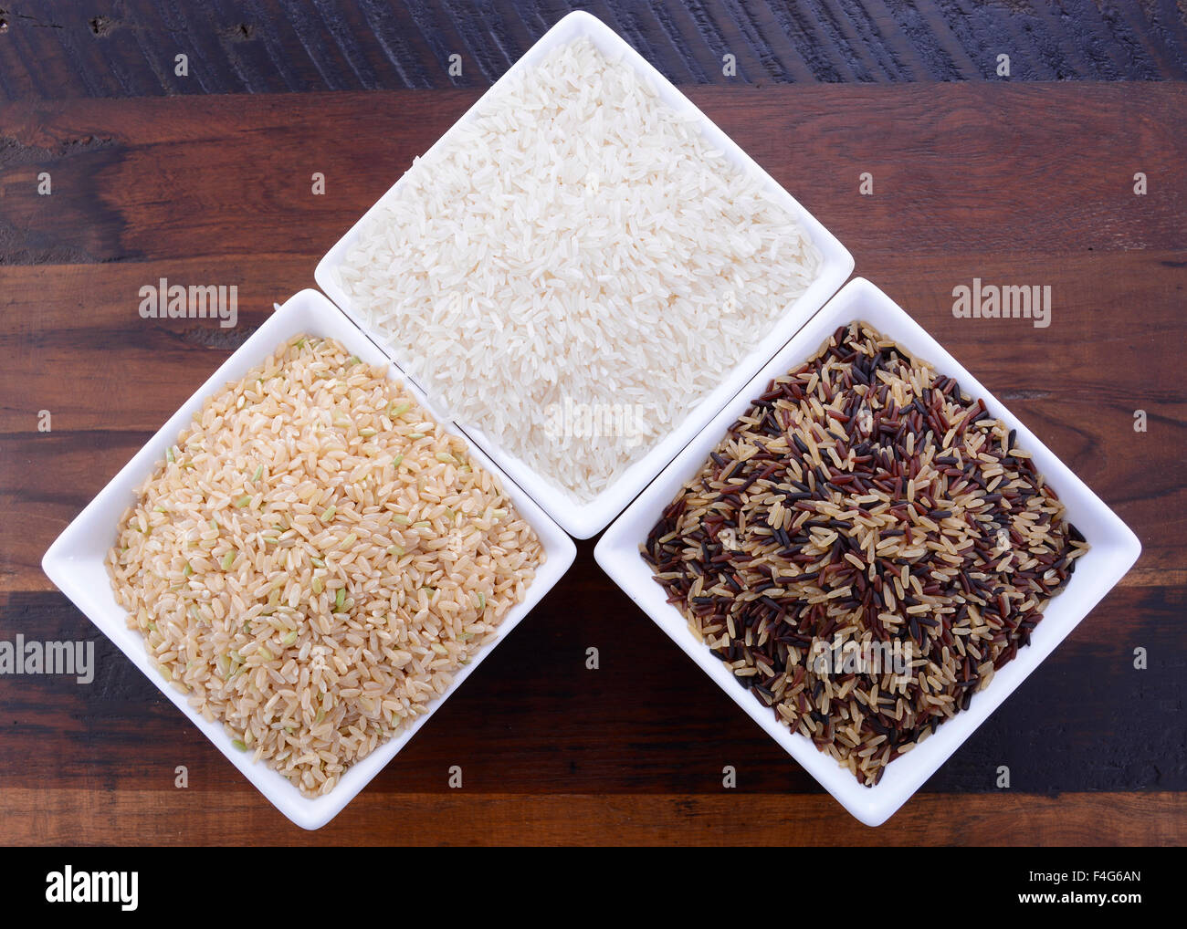 Square bowls of uncooked brown, white, and red and black rice on dark ...