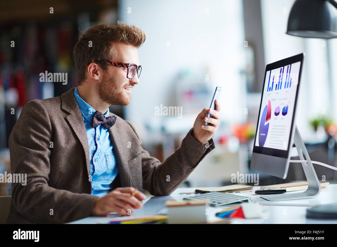 Young employee with cellphone sitting in front of computer Stock Photo ...