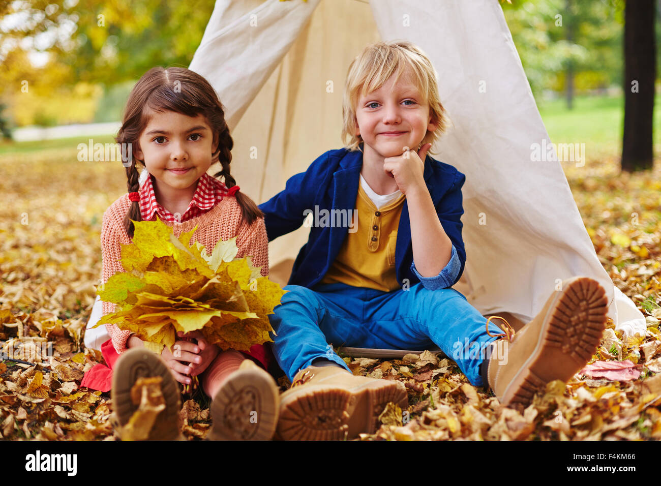 Two kids looking at camera while sitting on the ground in park in ...