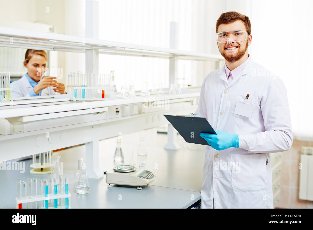 Happy chemist in white-coat, eyeglasses and gloves making notes in lab ...