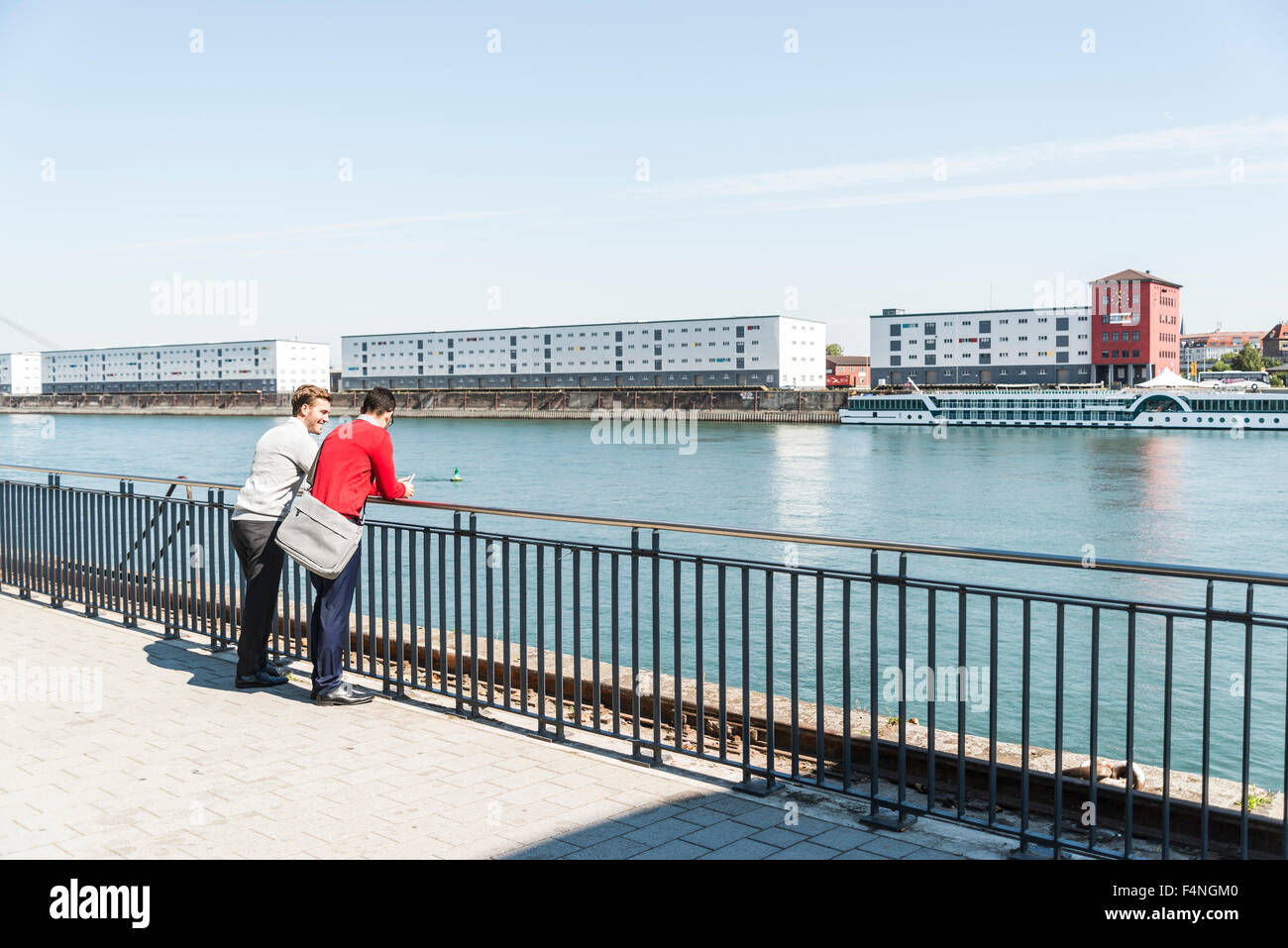 Two young businessmen standing by the riverside, leaning on railing ...