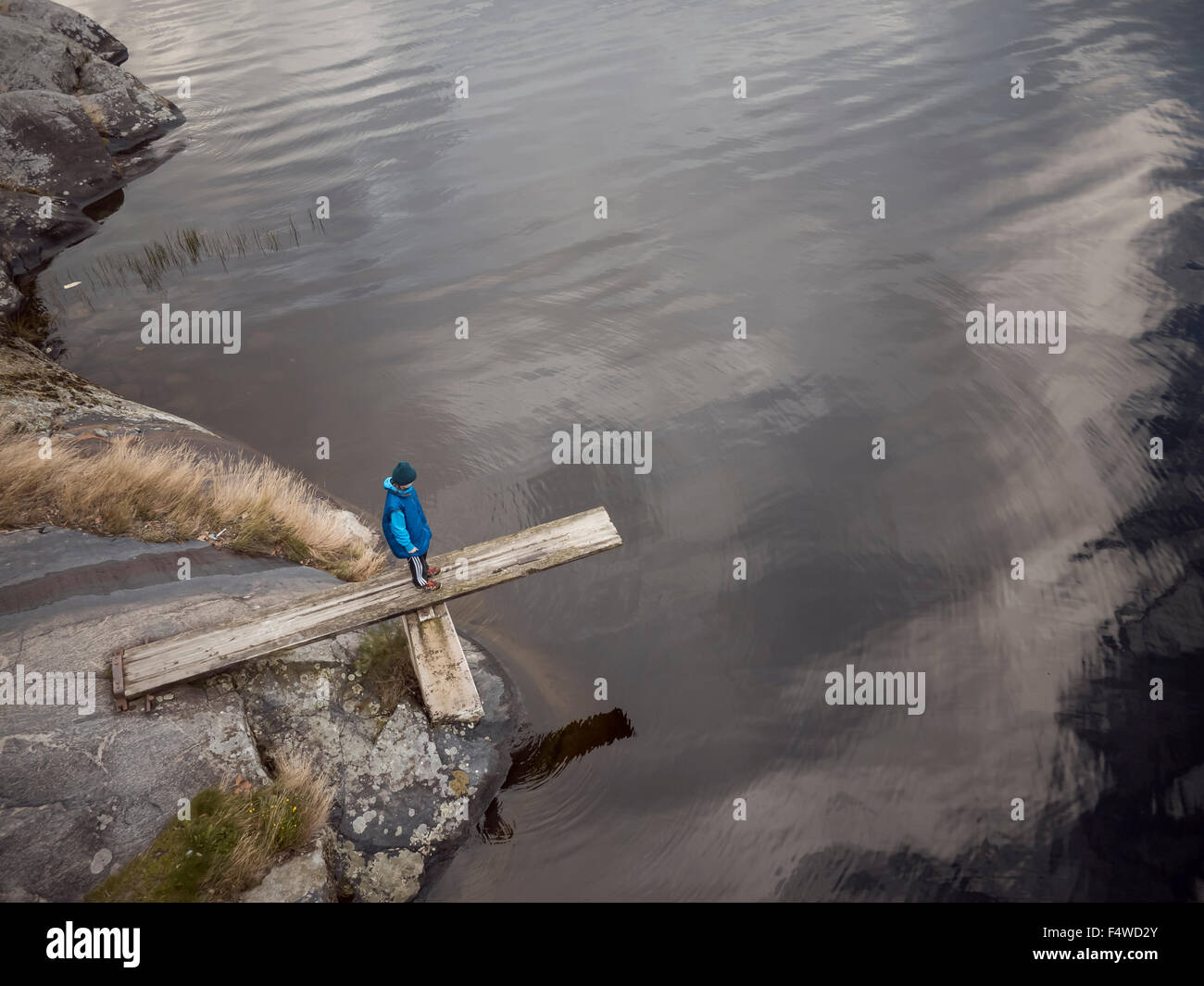 Boy (10-11) standing on springboard projecting off rocky lakeshore ...