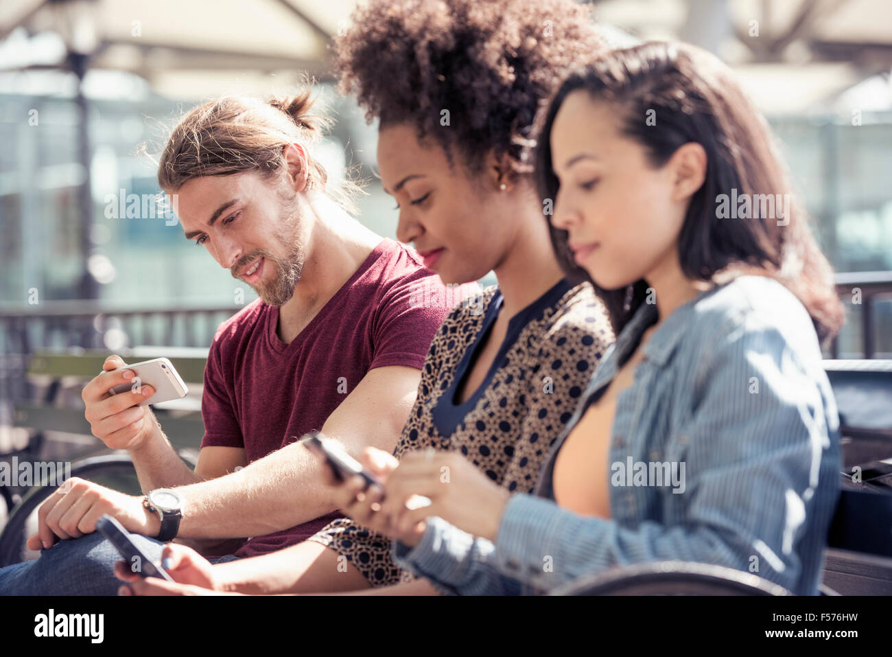 Three people seated side by side on a park bench checking their smart ...
