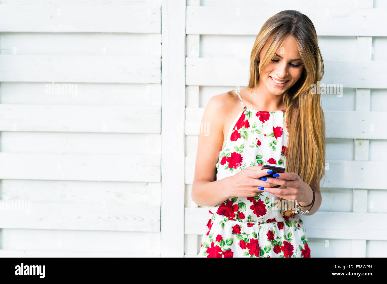 Beautiful young woman using phone while leaning against a white wall ...