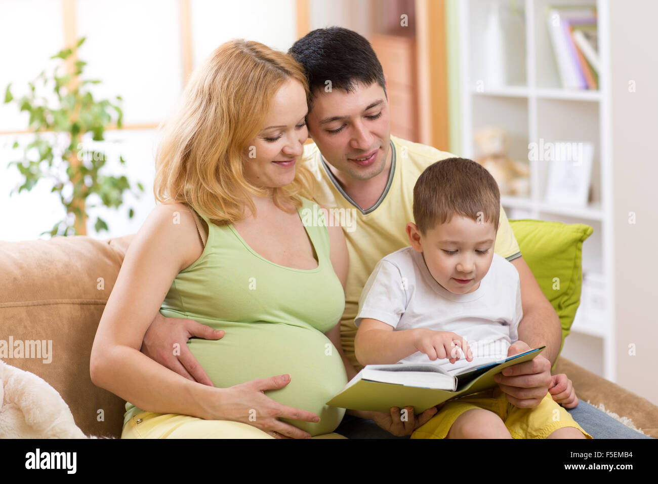 Happy parents reading book to child together on couch in home Stock ...