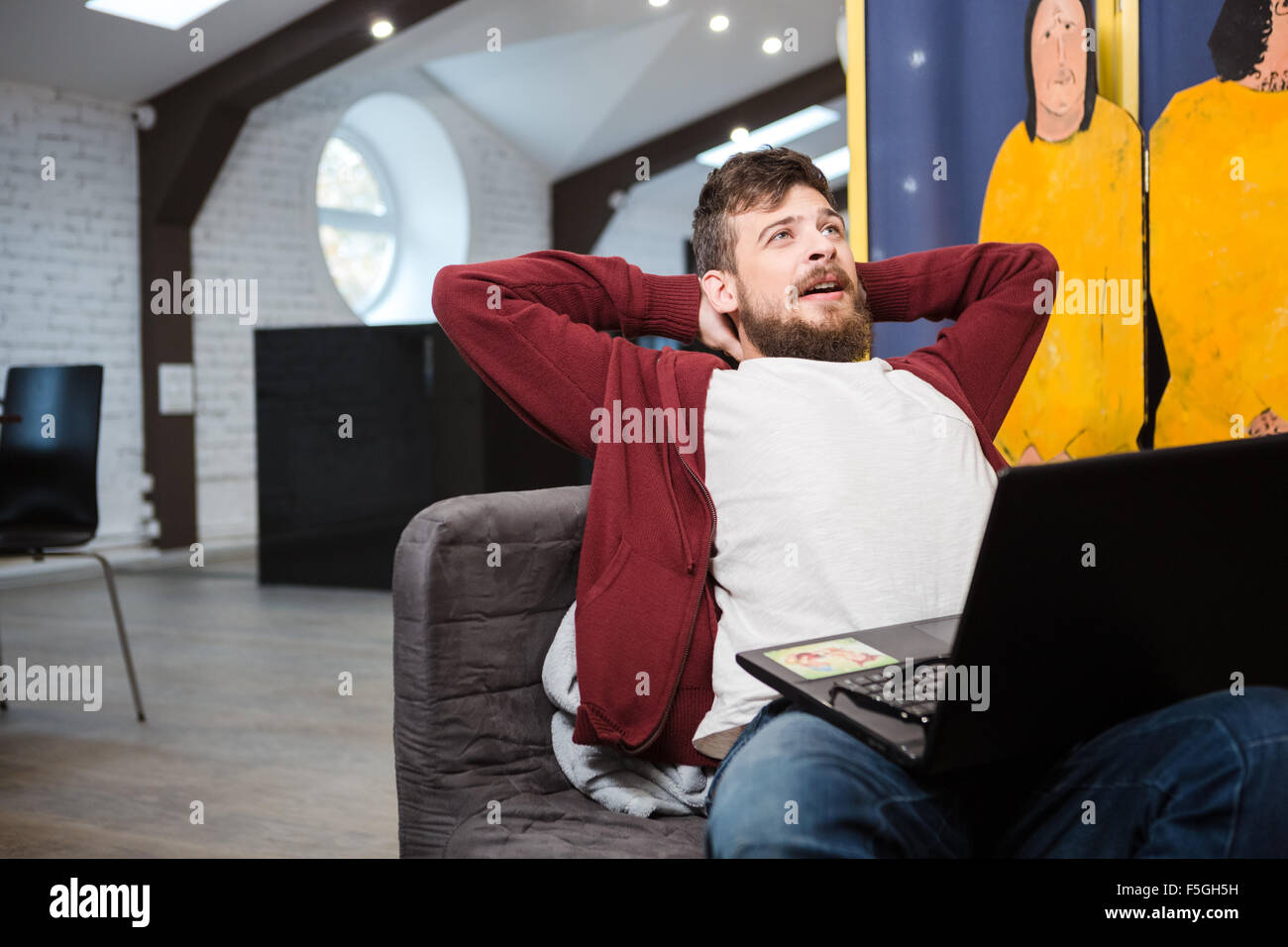 Handsome tired guy with beard yawning while working on laptop at office ...