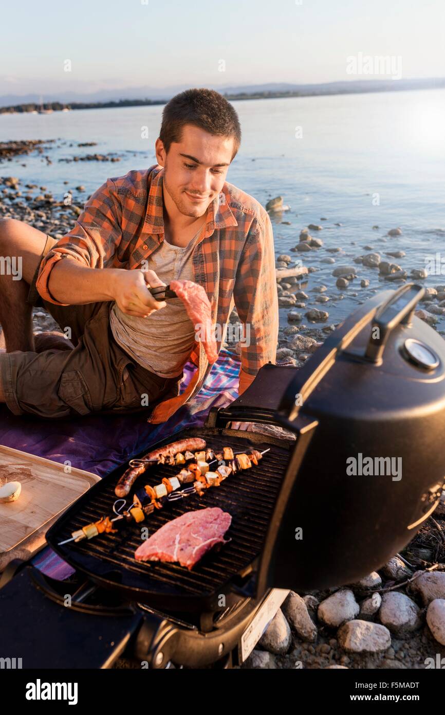 Young man sitting next to water cooking meat and kebabs on barbecue ...