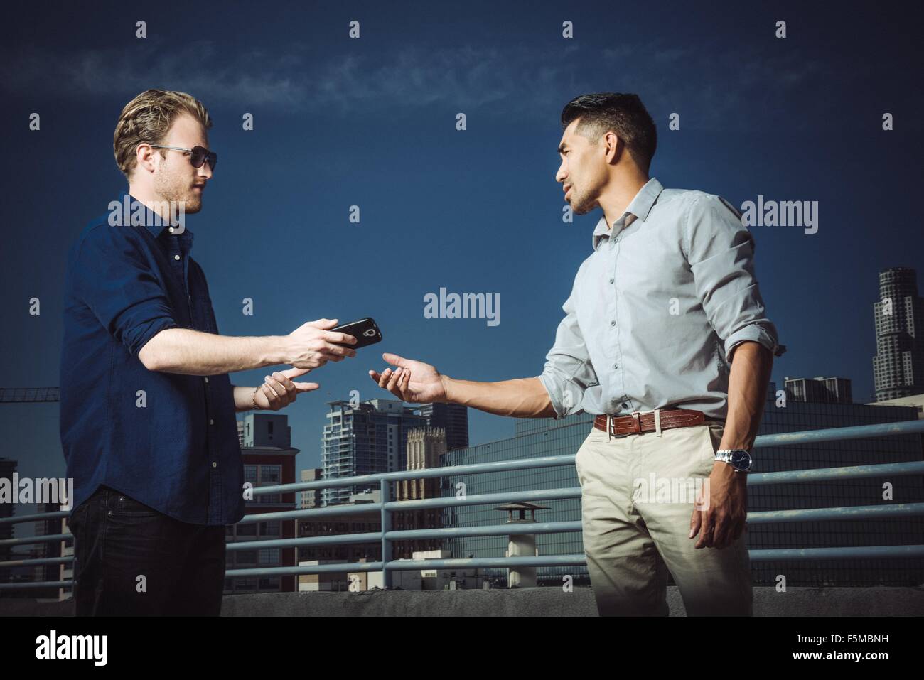 Businessman giving smartphone to man on roof terrace, Los Angeles ...
