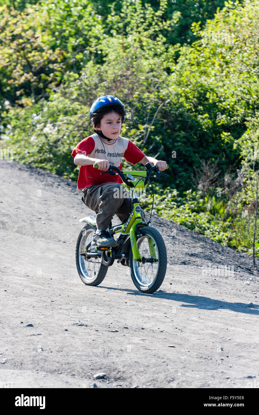 Caucasian 9 year old child, boy, riding green bike down sloping dirt track with bushes in ...