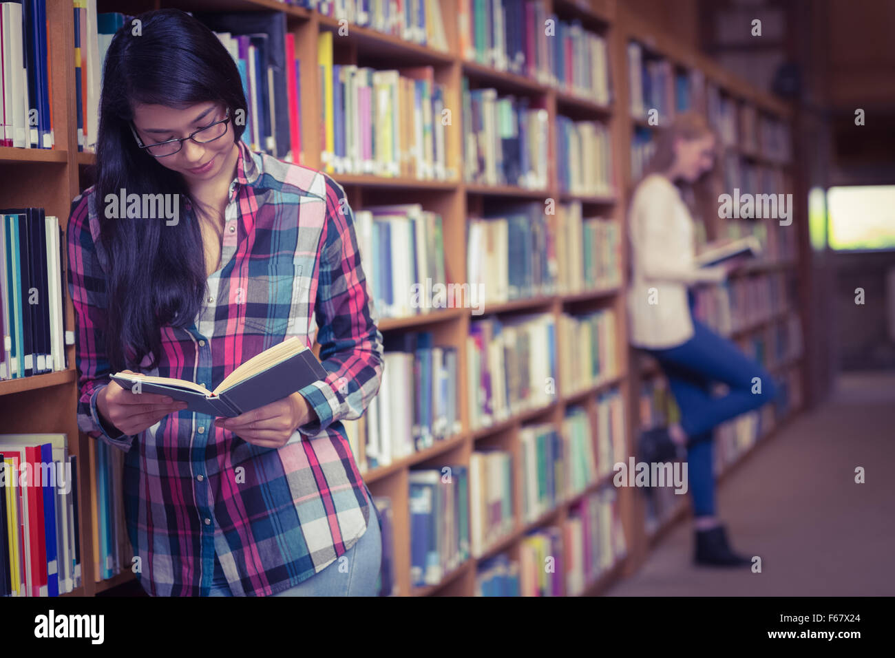 Smiling student leaning against bookshelves reading book Stock Photo ...