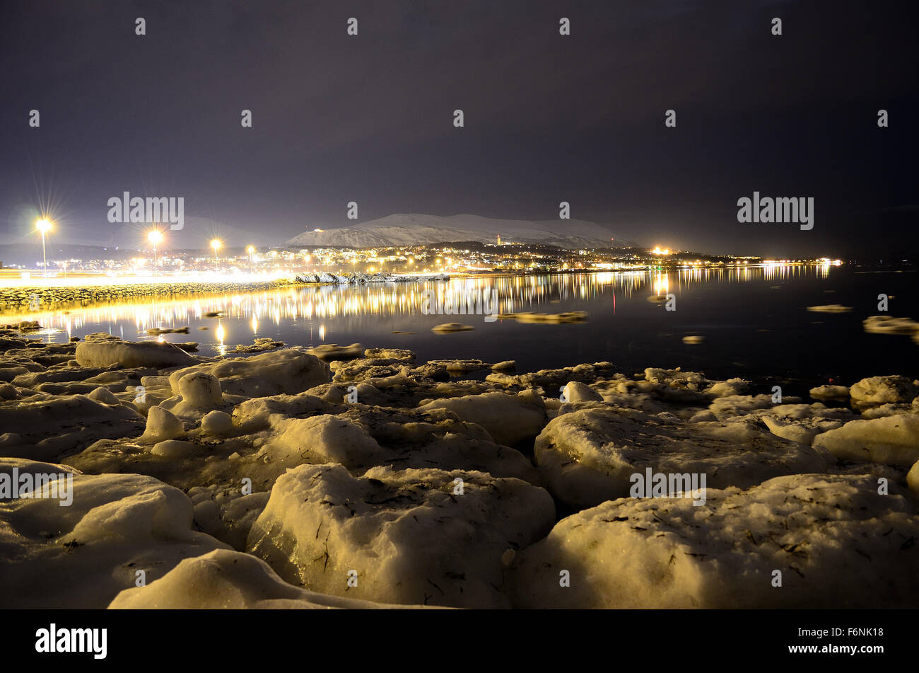 snowy sea shore with bright city lights and reflective fjord surface in ...