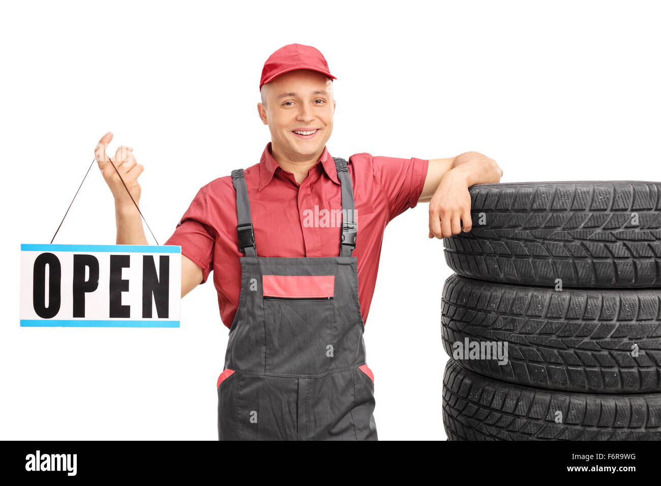 Young mechanic holding an open sign and leaning on a stack of car tires ...