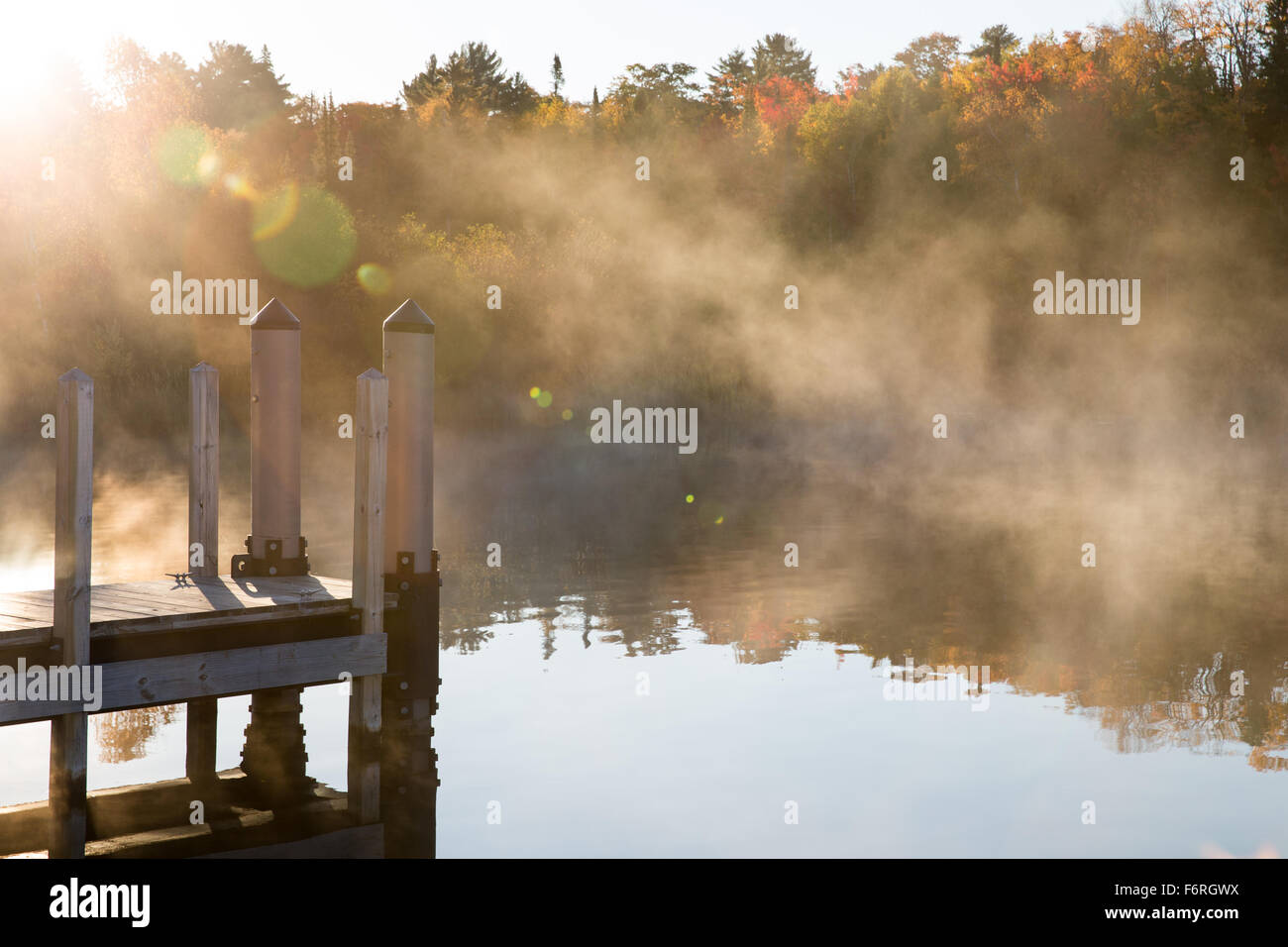 Morning fog hanging over a calm lake with sun and lens flare and a pier ...
