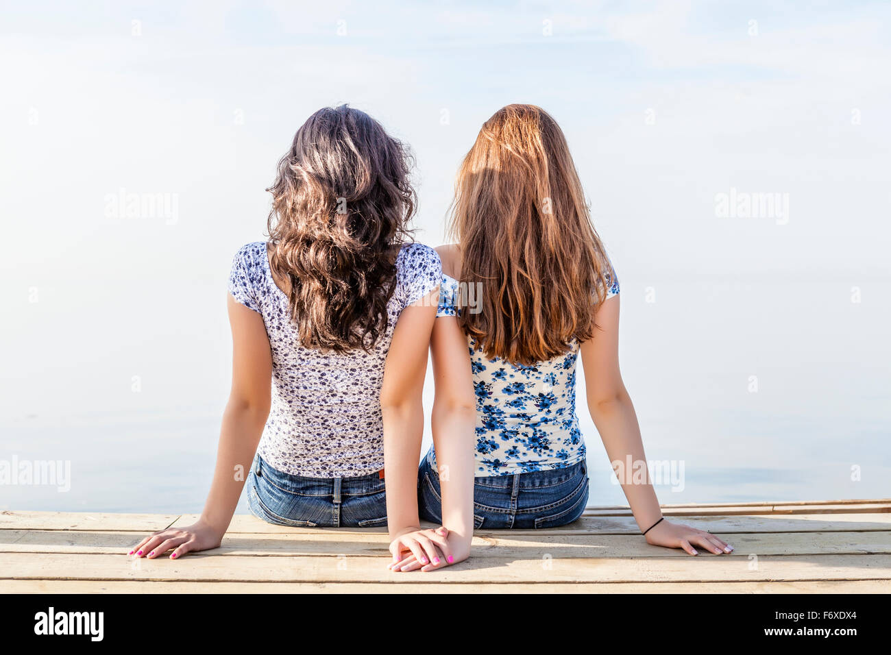 Two girls holding hands sitting close together at end of a dock ...