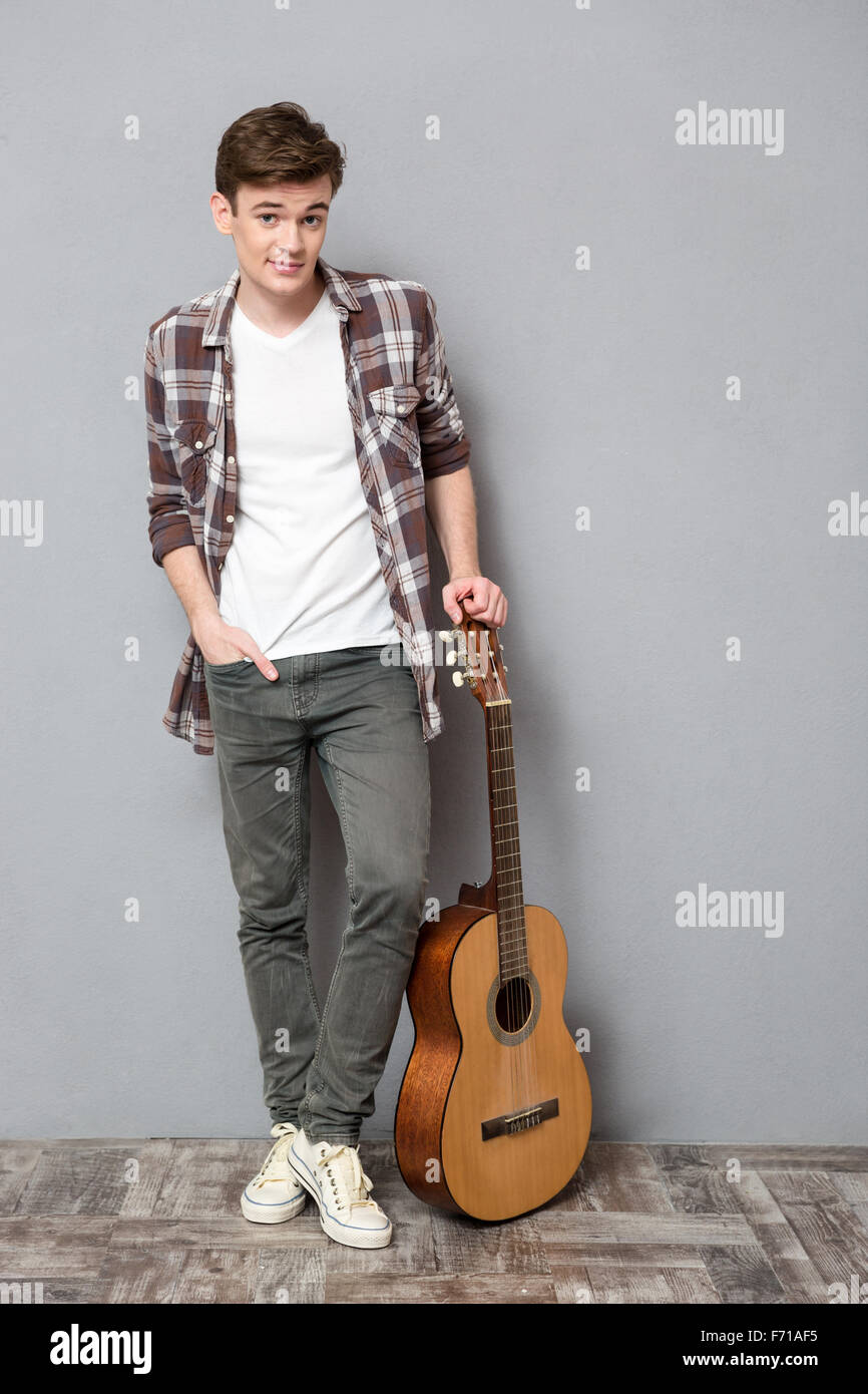 Full length portrait of a young man standing with guitar on gray ...