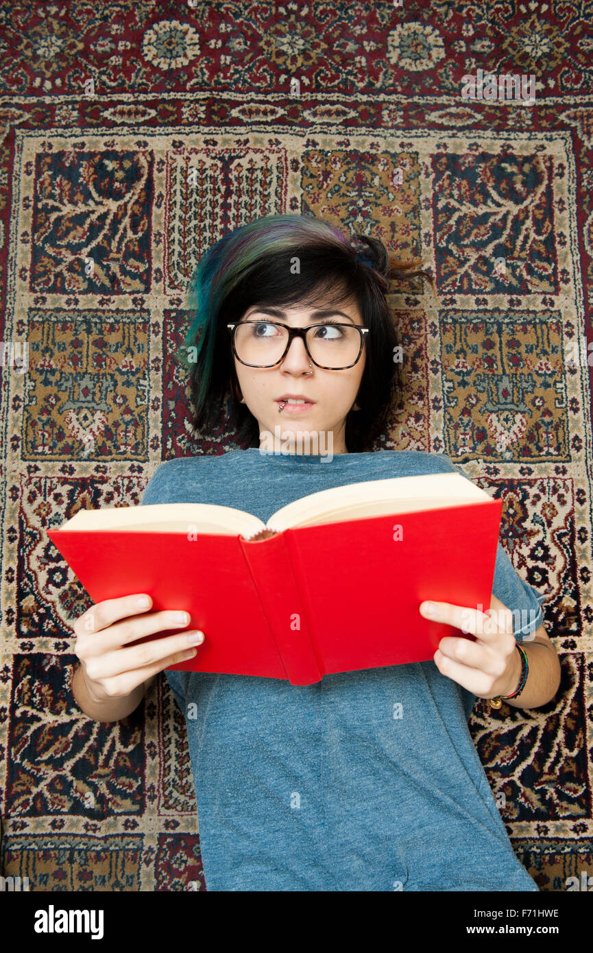 Pretty young woman lying on carpet and reading a red book while looking ...