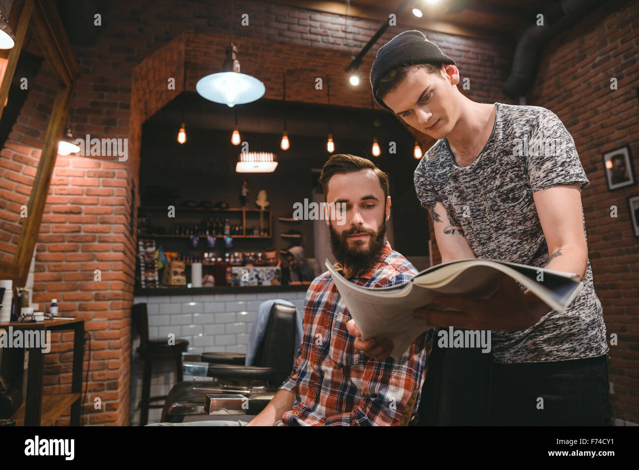 Young hairstylist and attractive bearded client looking through ...