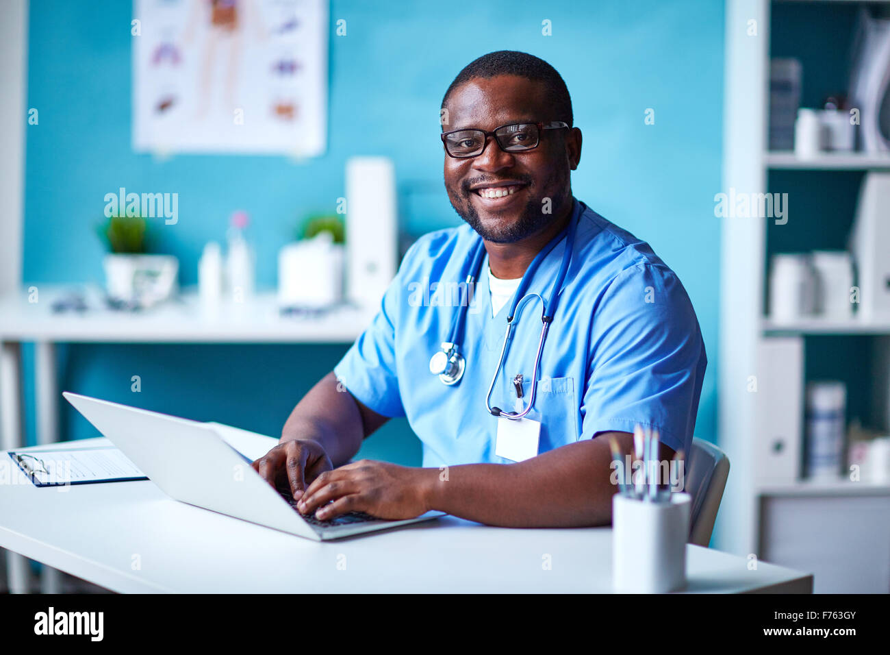 Modern doctor in uniform looking at camera while typing at workplace ...