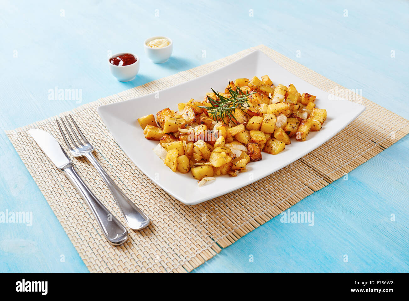 Roasted potatoes on square white plate, fork, knife, sauces and blue ...