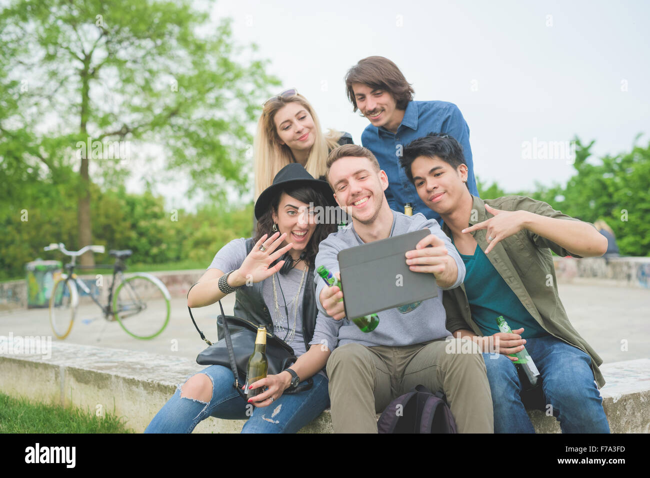 Group of young multiethnic friends sitting on a small wall using a ...