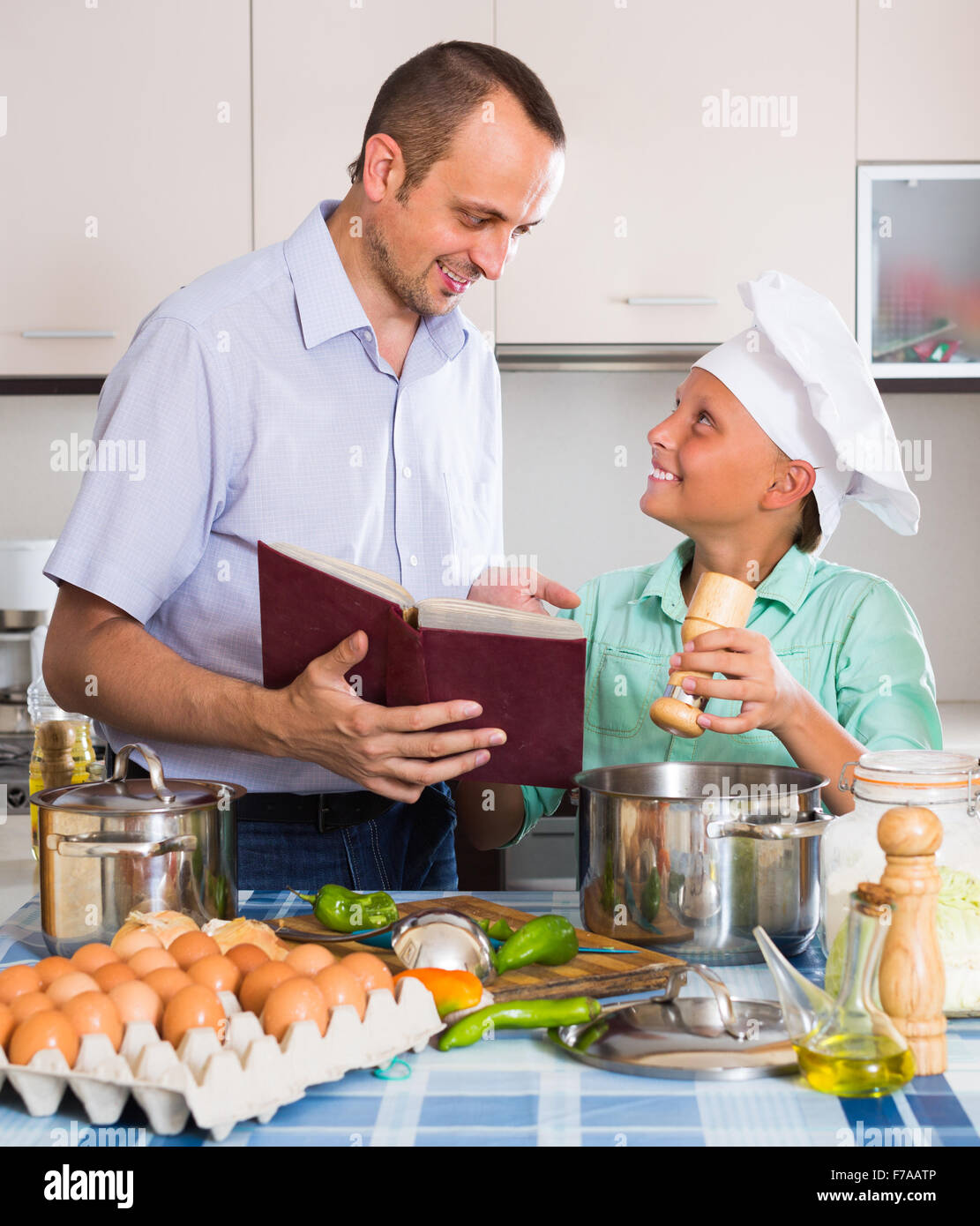 Joyful man and his teenage son cooking together with recipe book at ...