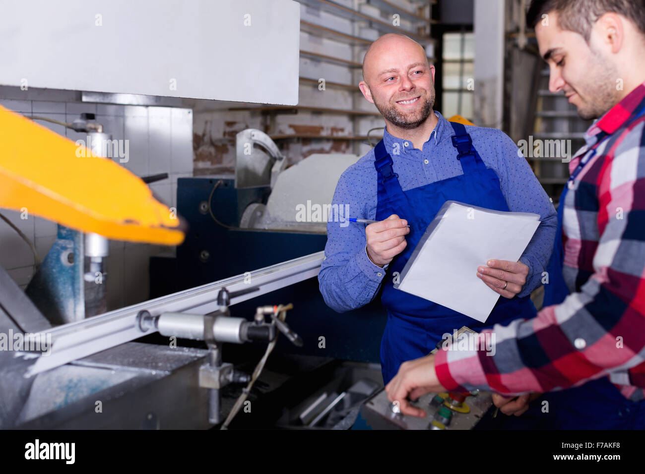 Smiling worker in coverall operating in lathe, his boss with papers ...