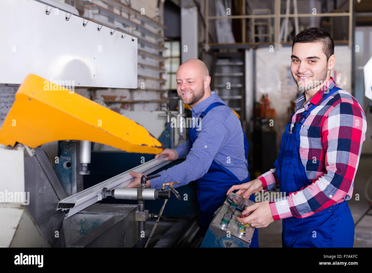 Two cheerful workers in uniform working on a machine in PVC shop and ...