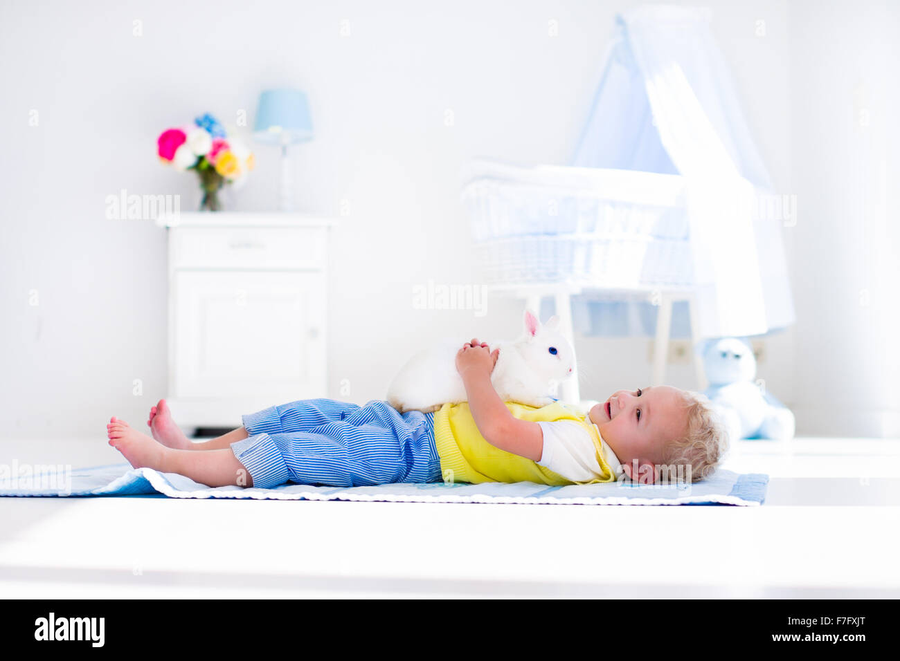 Cute blond curly baby boy playing with rabbit in a white sunny bedroom ...