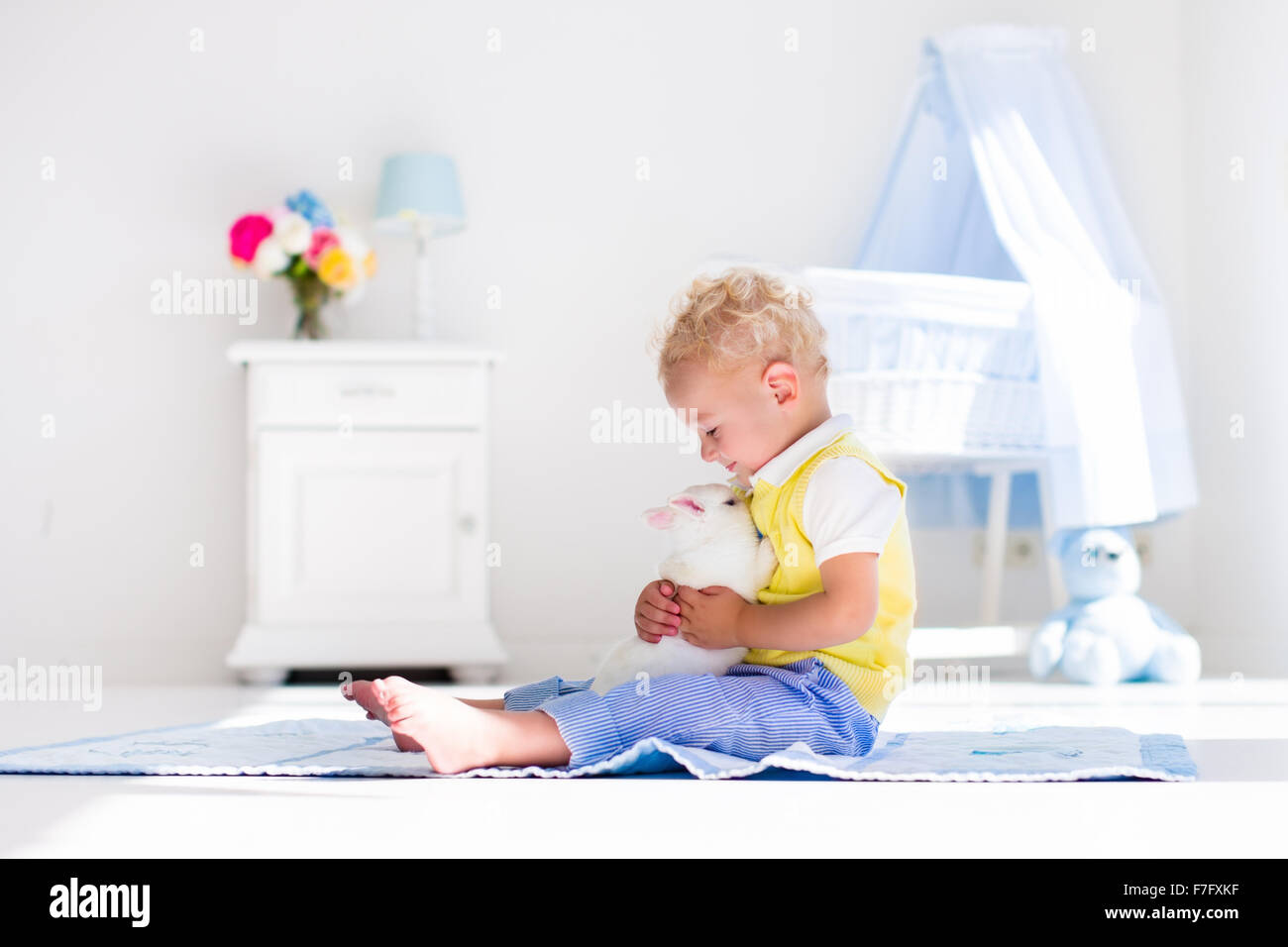Cute blond curly baby boy playing with rabbit in a white sunny bedroom ...