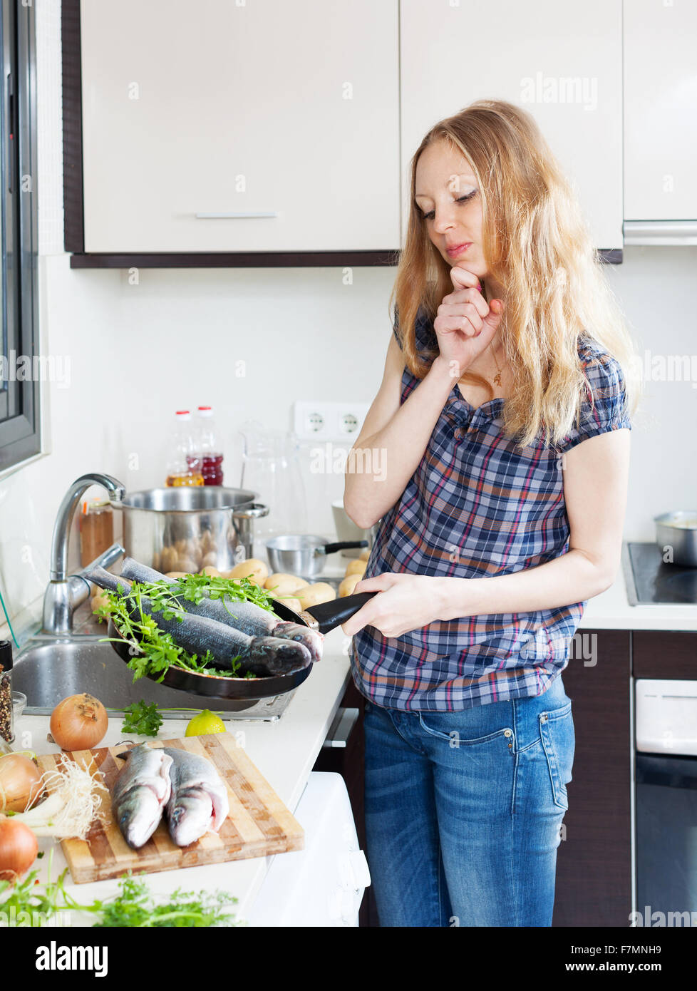 Long-haired woman is thinking how to cook fish in fryingpan at kitchen ...