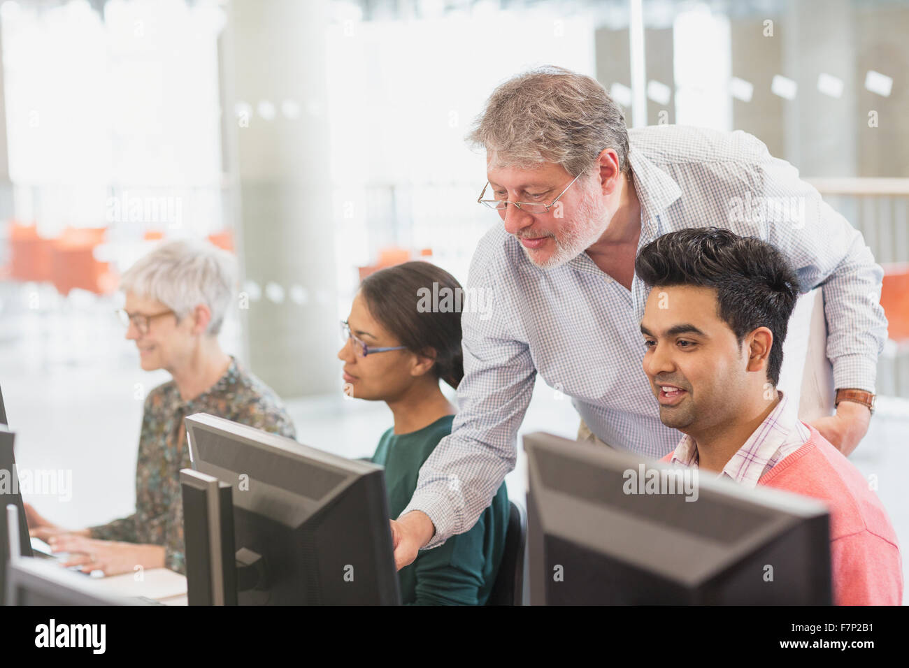 Professor helping student at computer in adult education classroom ...