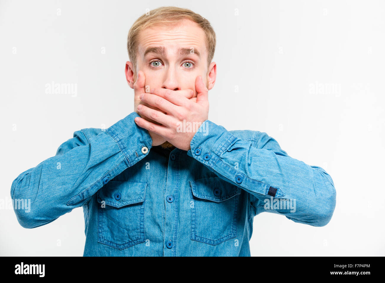 Shocked dazed young blond man closed his mouth by hands over white ...