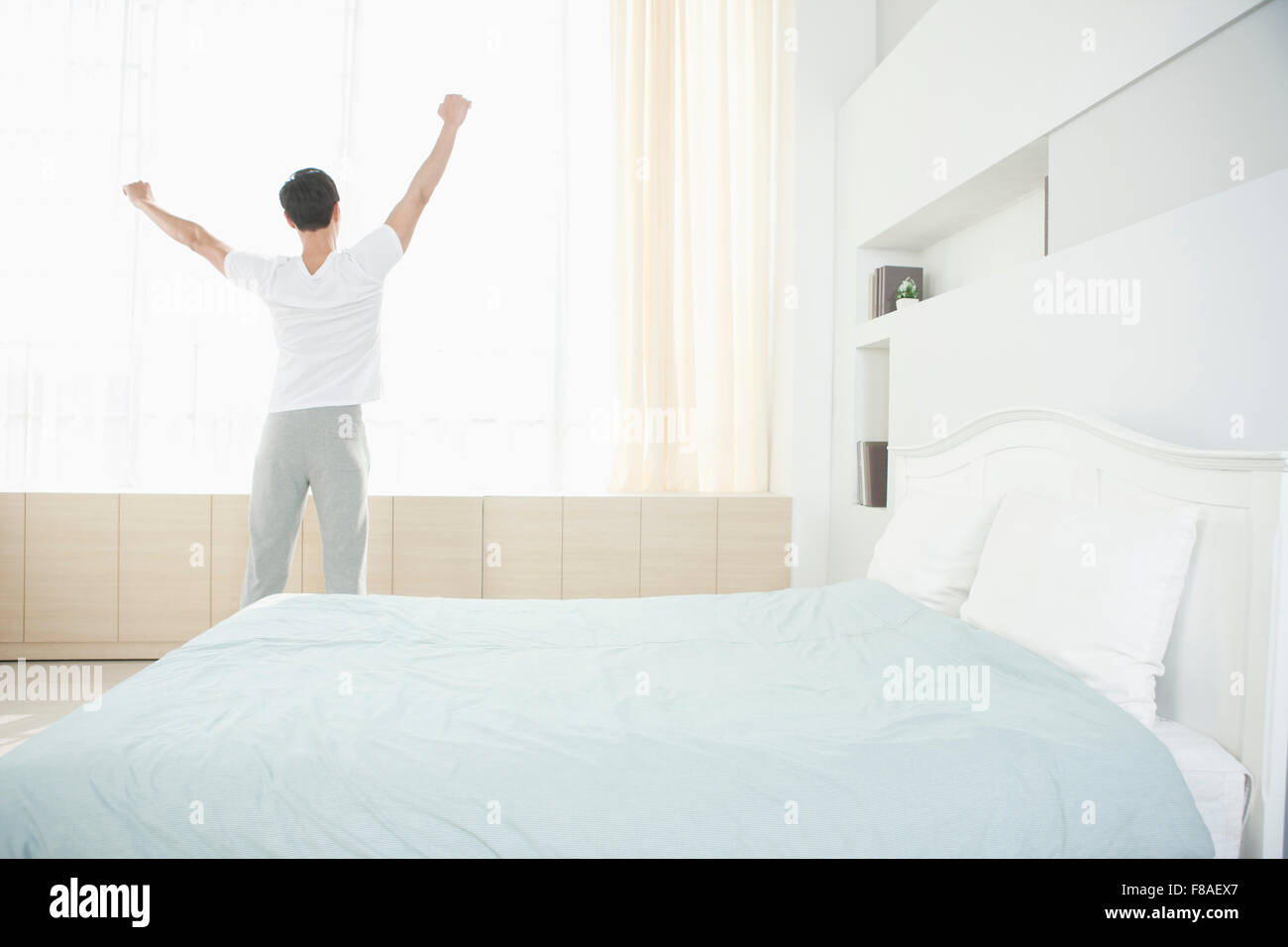 Asian man stretching his body next to the bed and looking out the ...