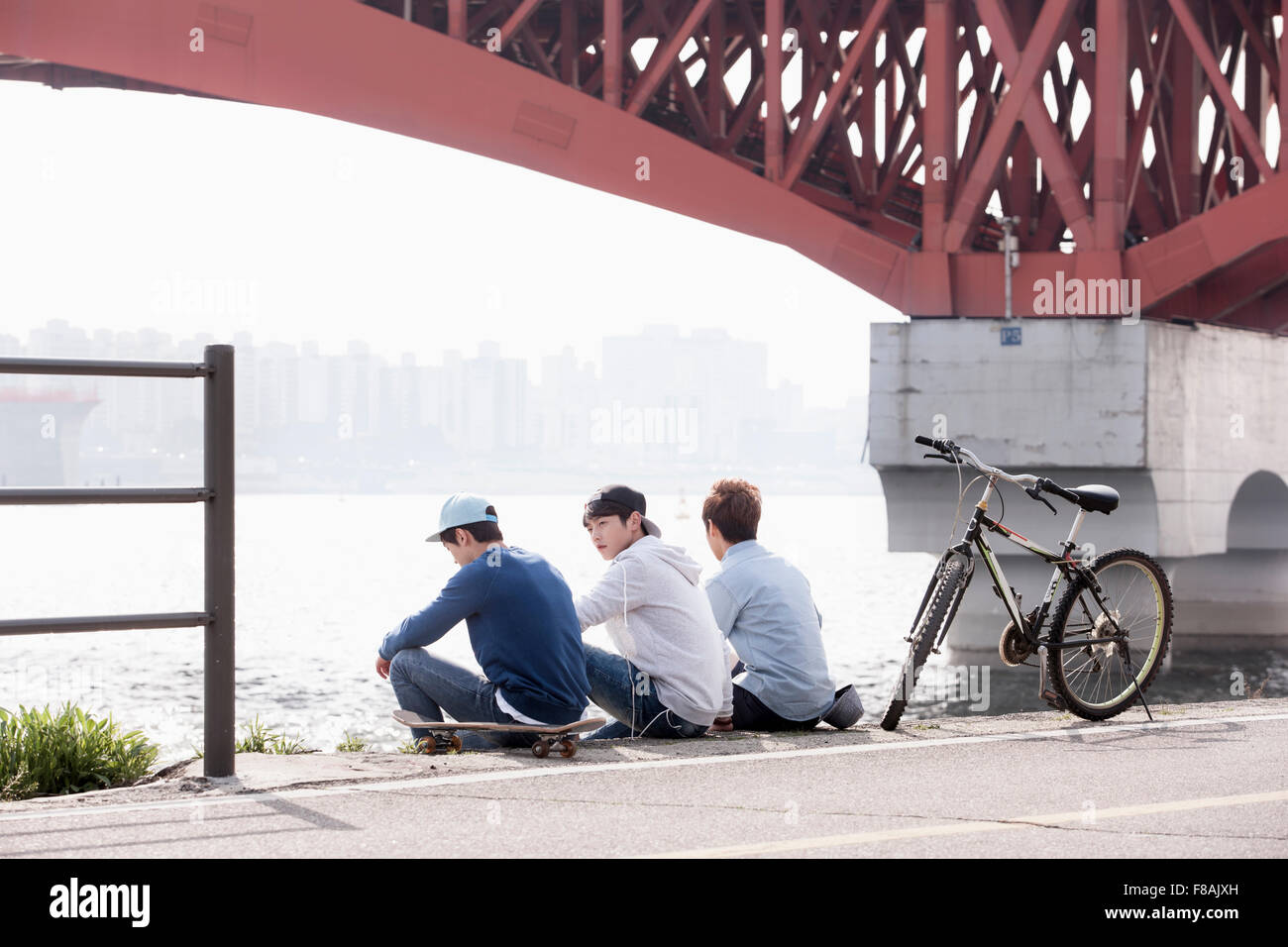 Back appearance of three young men sitting at the riverside under the ...