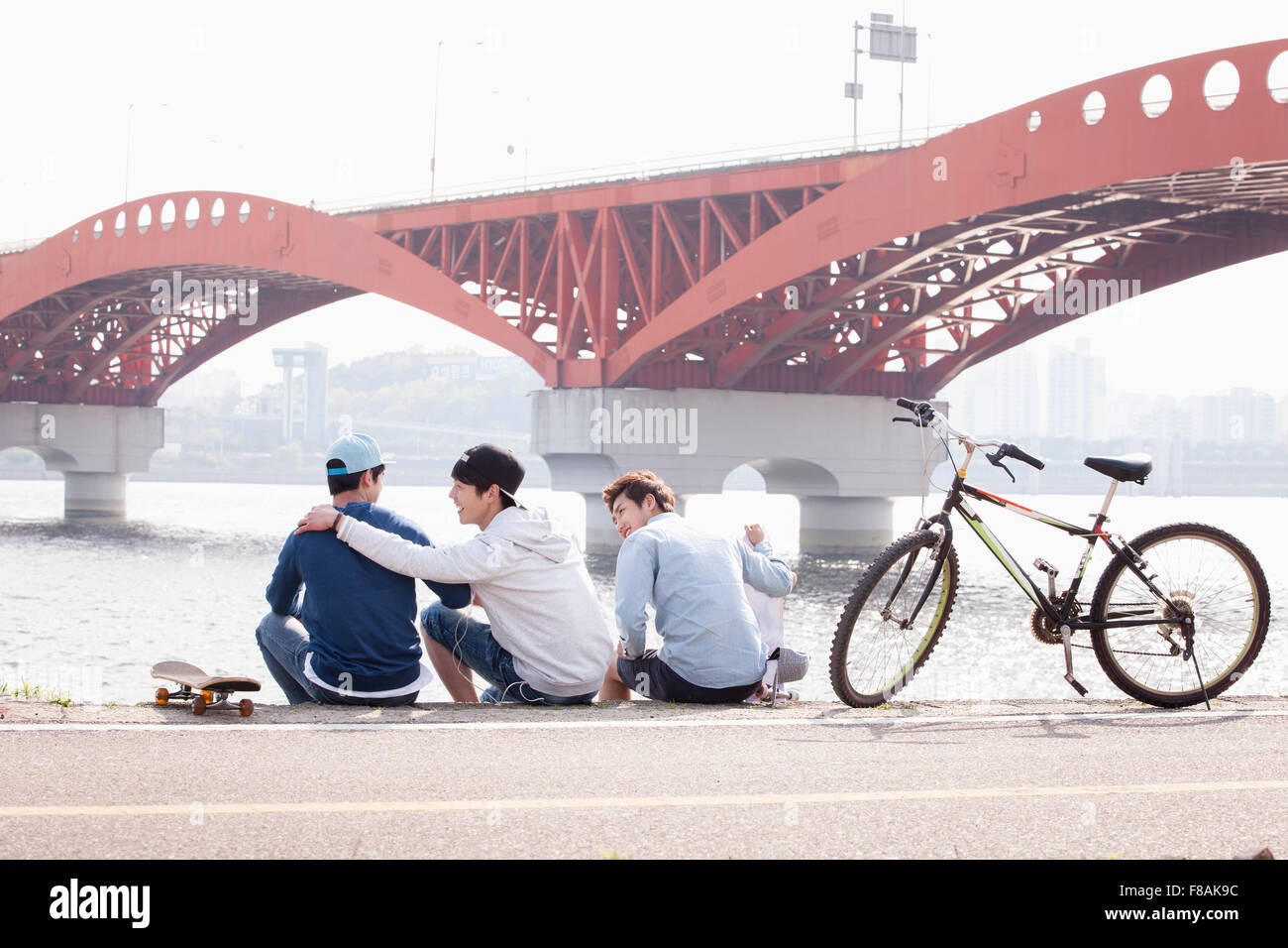 Three young men sitting at the riverside under the bridge and taking a ...