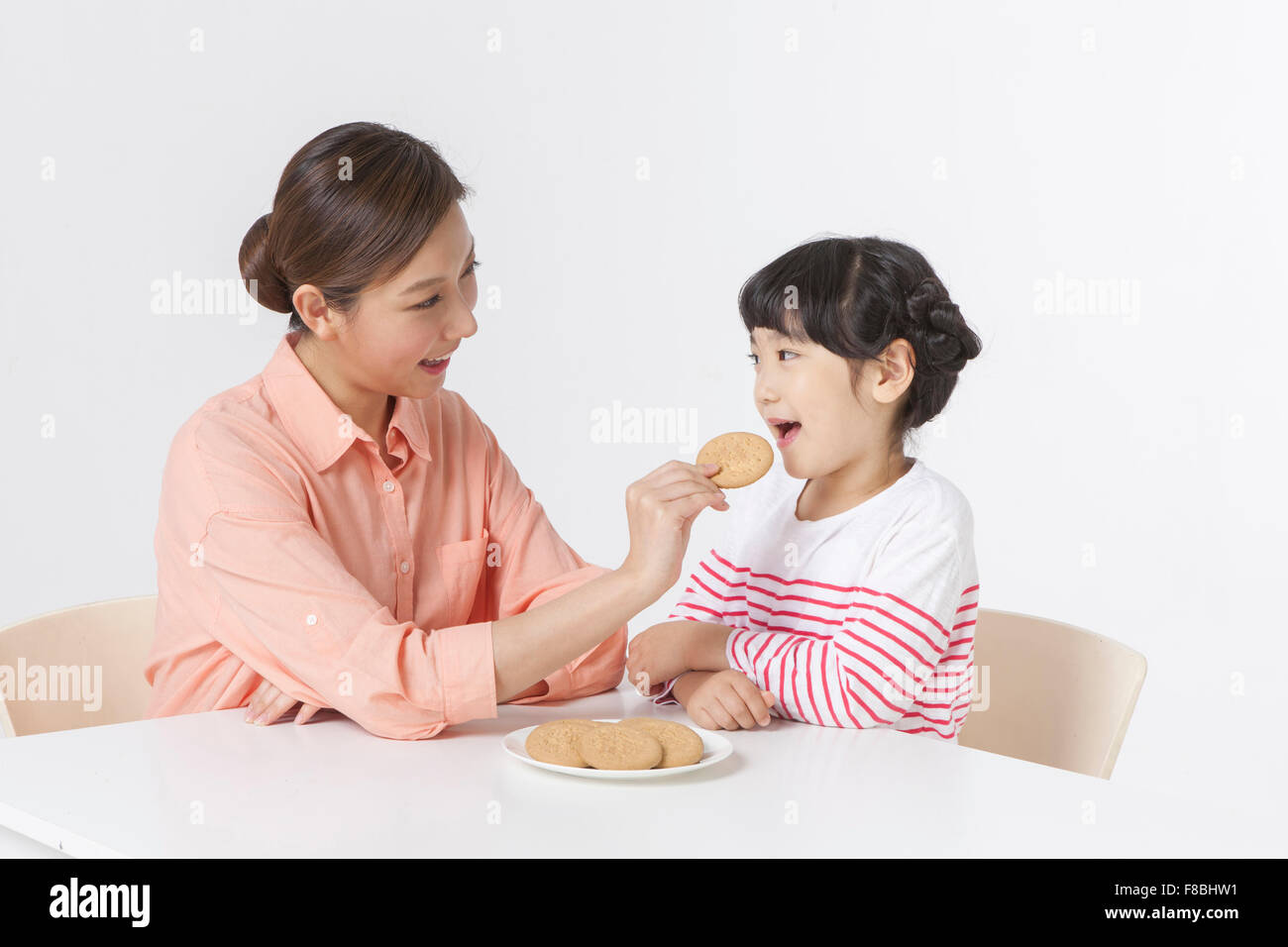 Mother giving a cookie to her daughter both seated at table Stock Photo ...