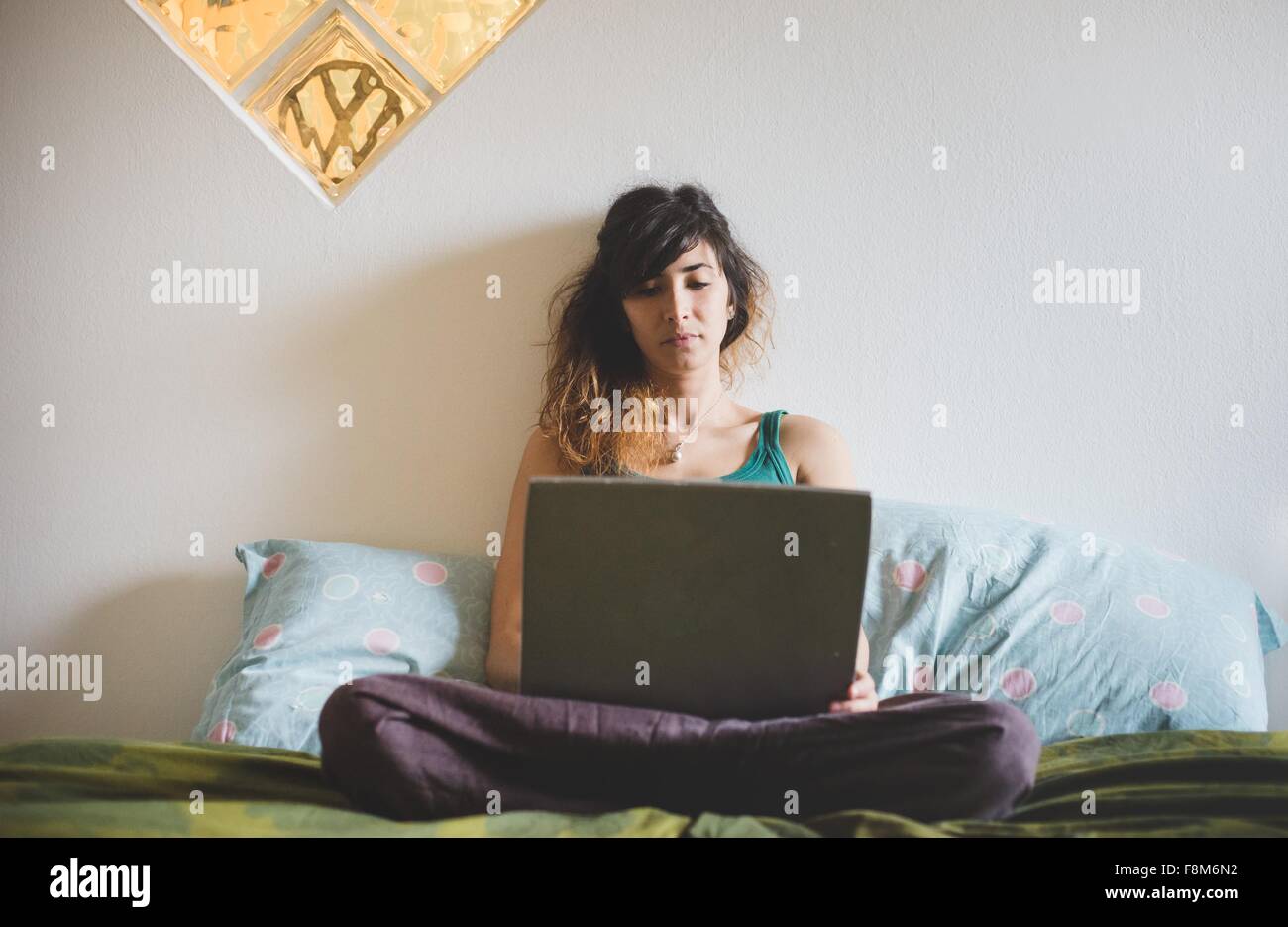 Front view of mid adult woman sitting cross legged on bed using laptop ...