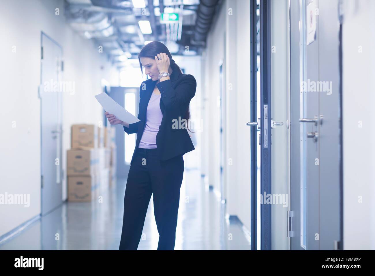 Young woman standing in office corridor looking down at paperwork ...